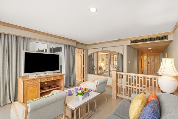 Quarto de hotel moderno com televisão de tela plana, área de refeições com mesa e cadeiras, sofá com almofadas estampadas e uma grande janela com cortinas.