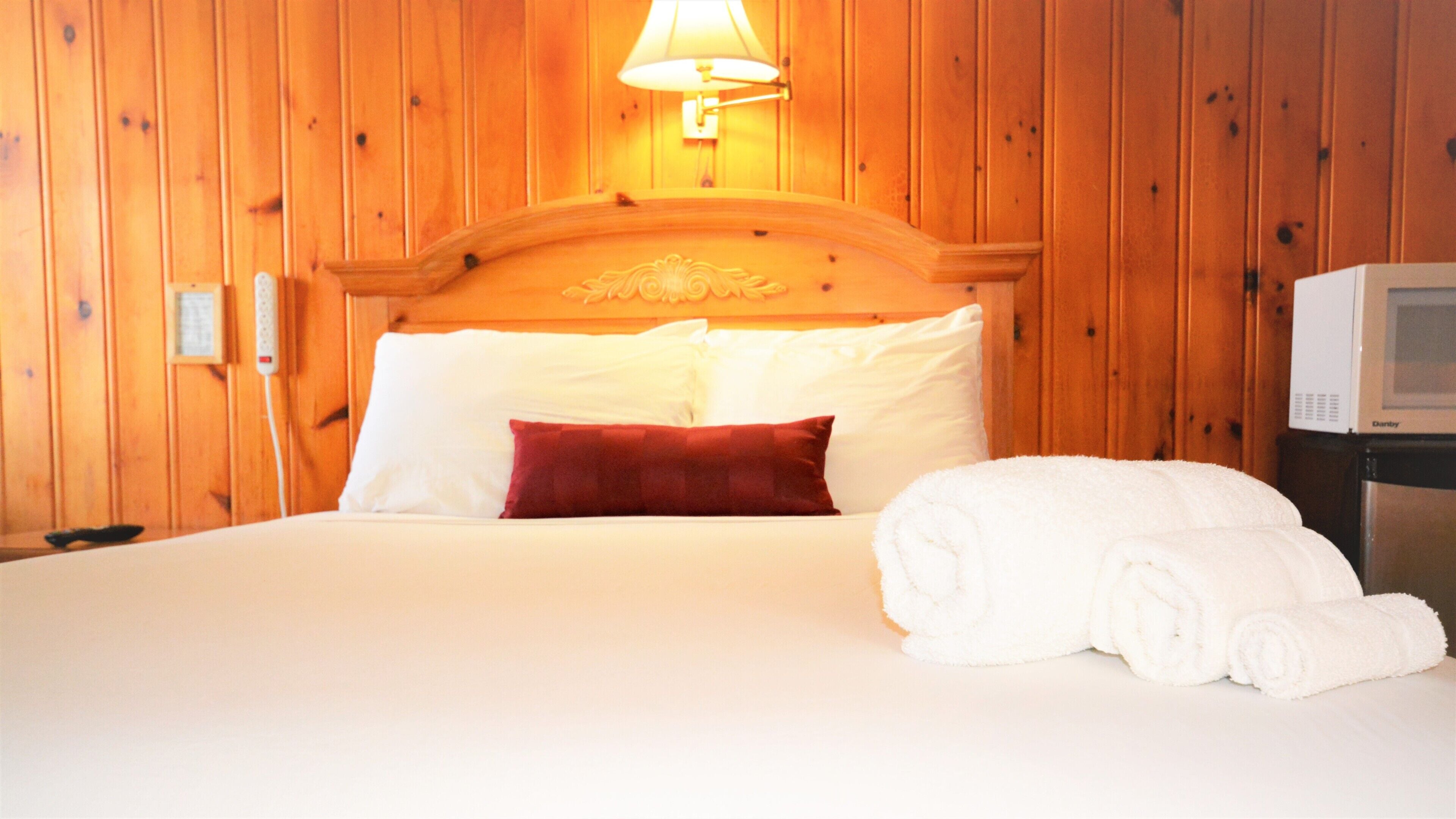 Quarto de hotel com cabeceira de madeira, roupa de cama branca, almofada vermelha e toalha branca enrolada na cama.