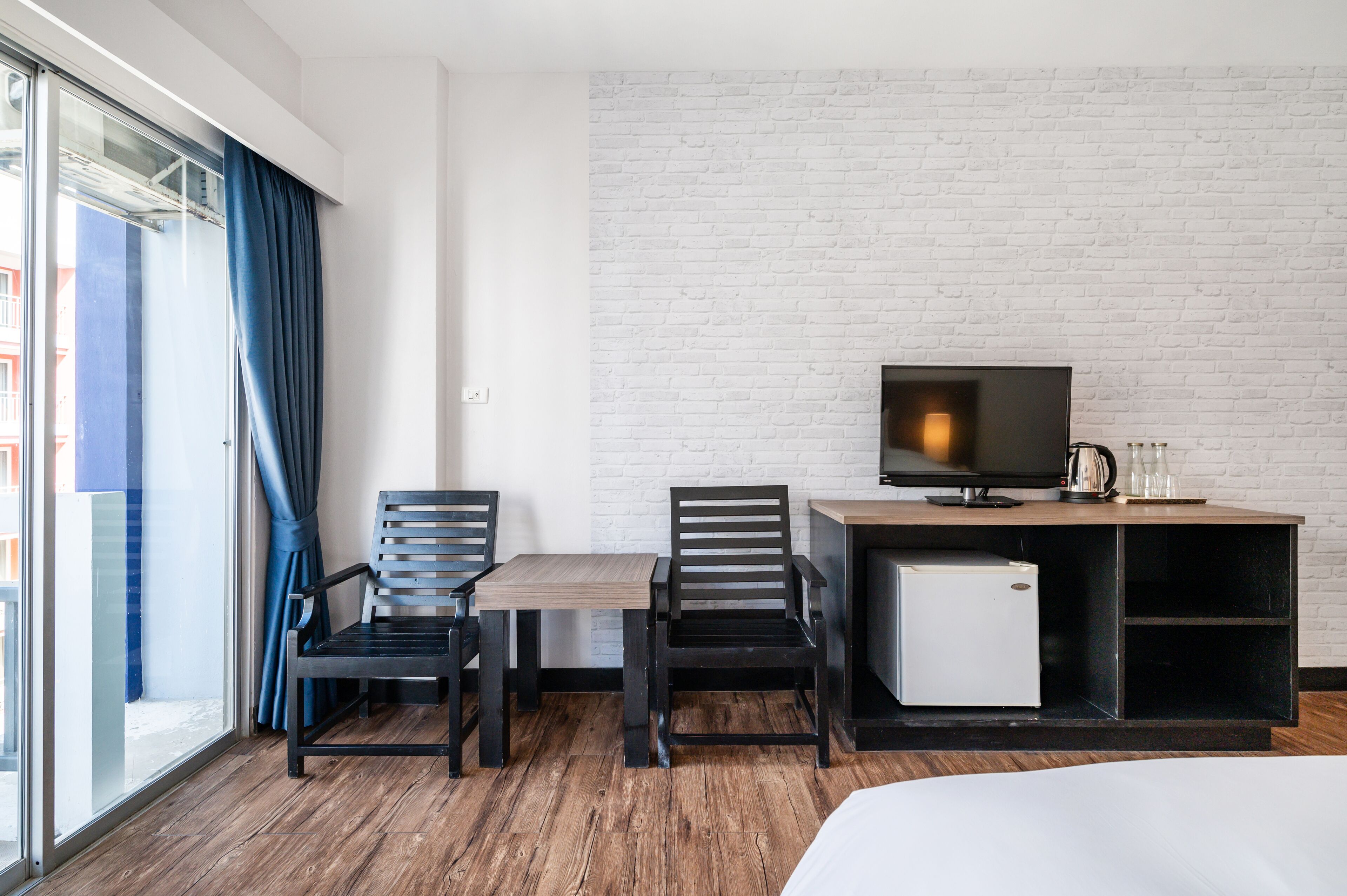 Quarto de hotel com piso de madeira, duas cadeiras pretas, uma geladeira branca pequena, uma TV sobre um suporte de madeira e uma parede de tijolos brancos.