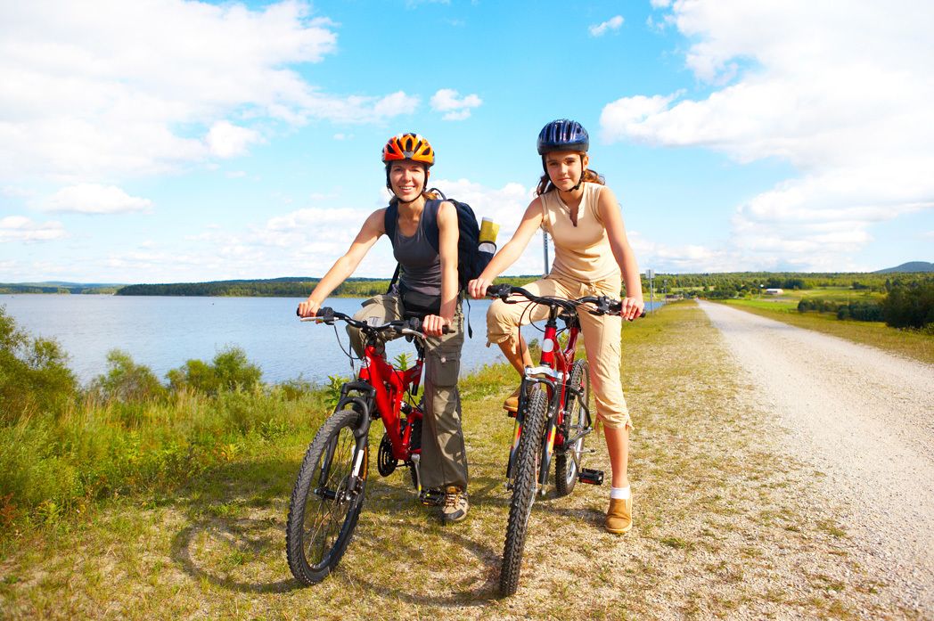 Duas pessoas de bicicleta perto de um lago, com céu claro.