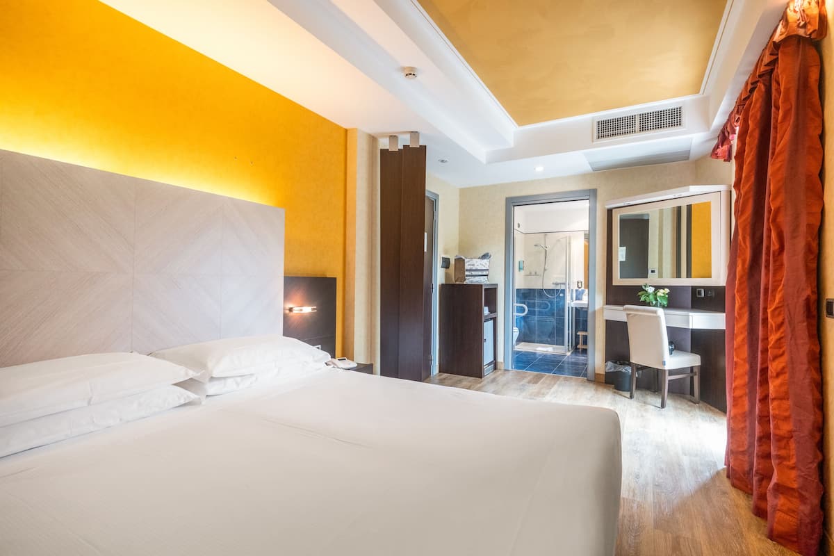 Quarto de hotel com uma cama grande, uma escrivaninha, um espelho e um banheiro visível por uma porta aberta.