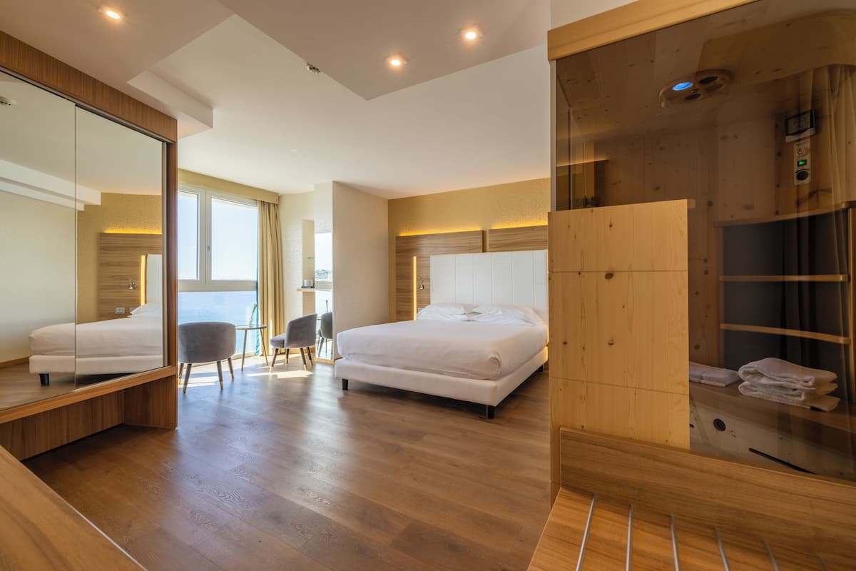 Quarto de hotel moderno com uma cama grande, um guarda-roupa de madeira, um espaço de estar com uma poltrona e um separador de vidro.