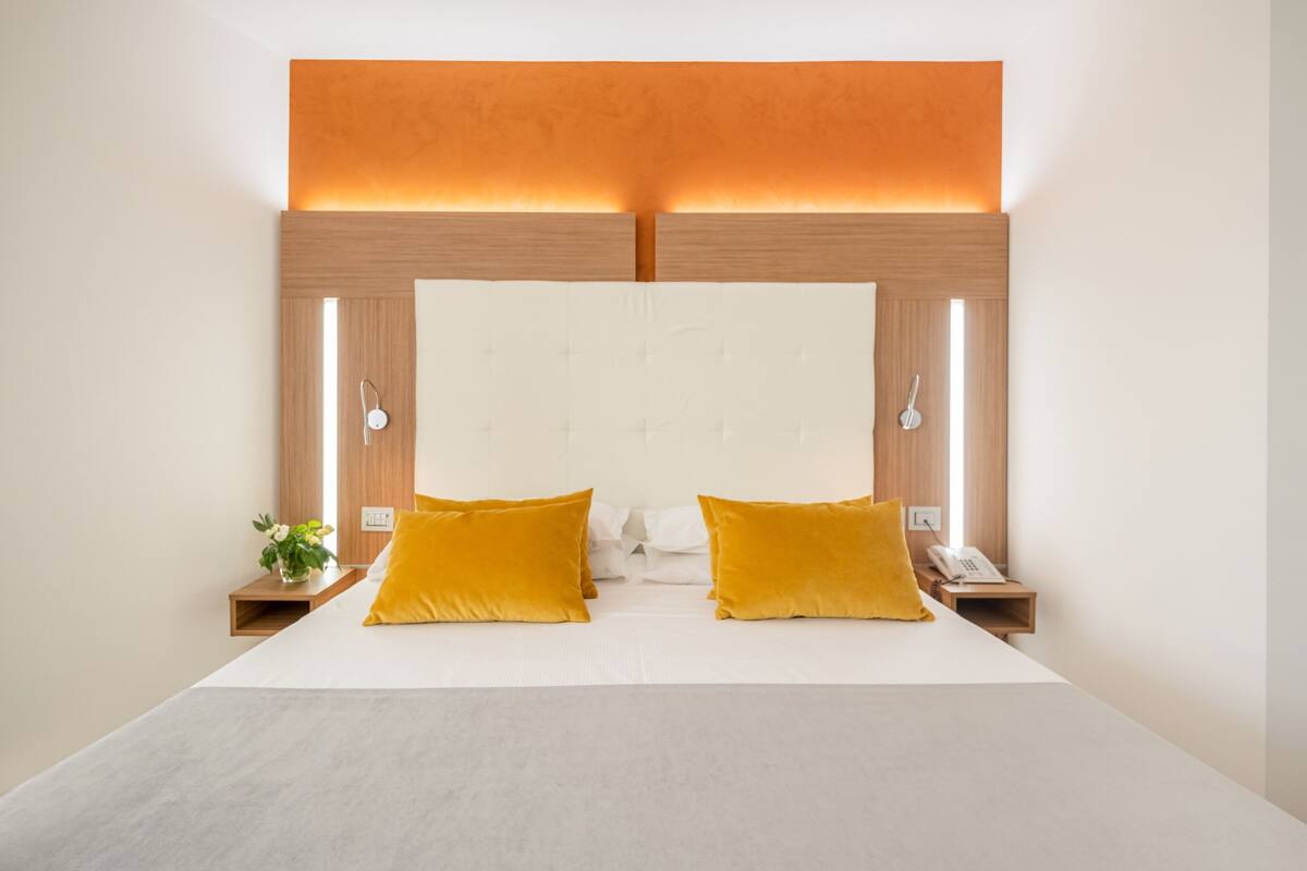 Quarto de hotel moderno com uma cama grande, duas mesas de cabeceira, um criado-mudo com telefone e um aplique na parede.
