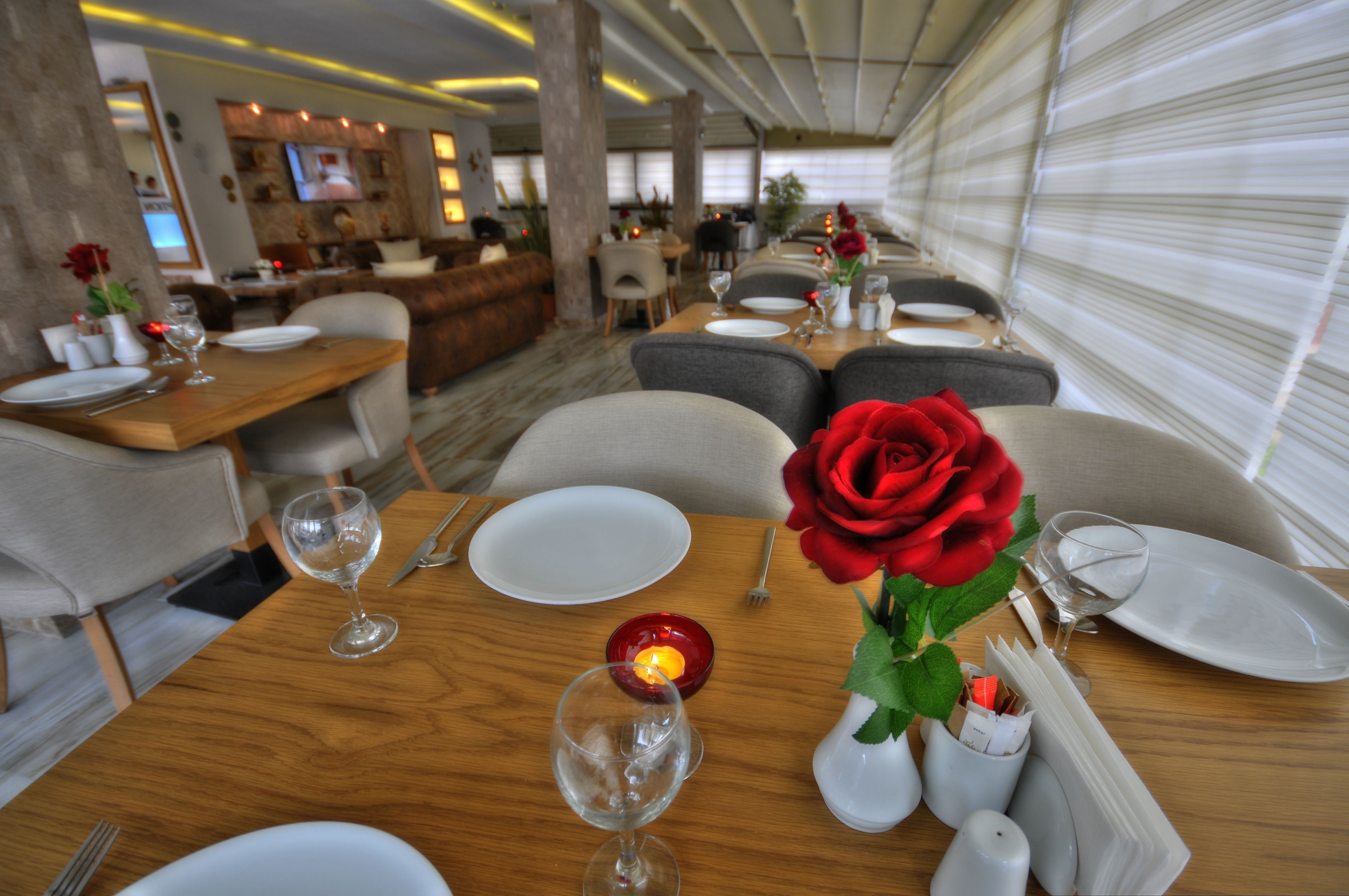 Uma sala de jantar com mesas preparadas para dois, com velas, copos de vinho e um arranjo central com uma rosa vermelha.