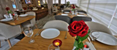 Uma sala de jantar com mesas preparadas para dois, com velas, copos de vinho e um arranjo central com uma rosa vermelha.