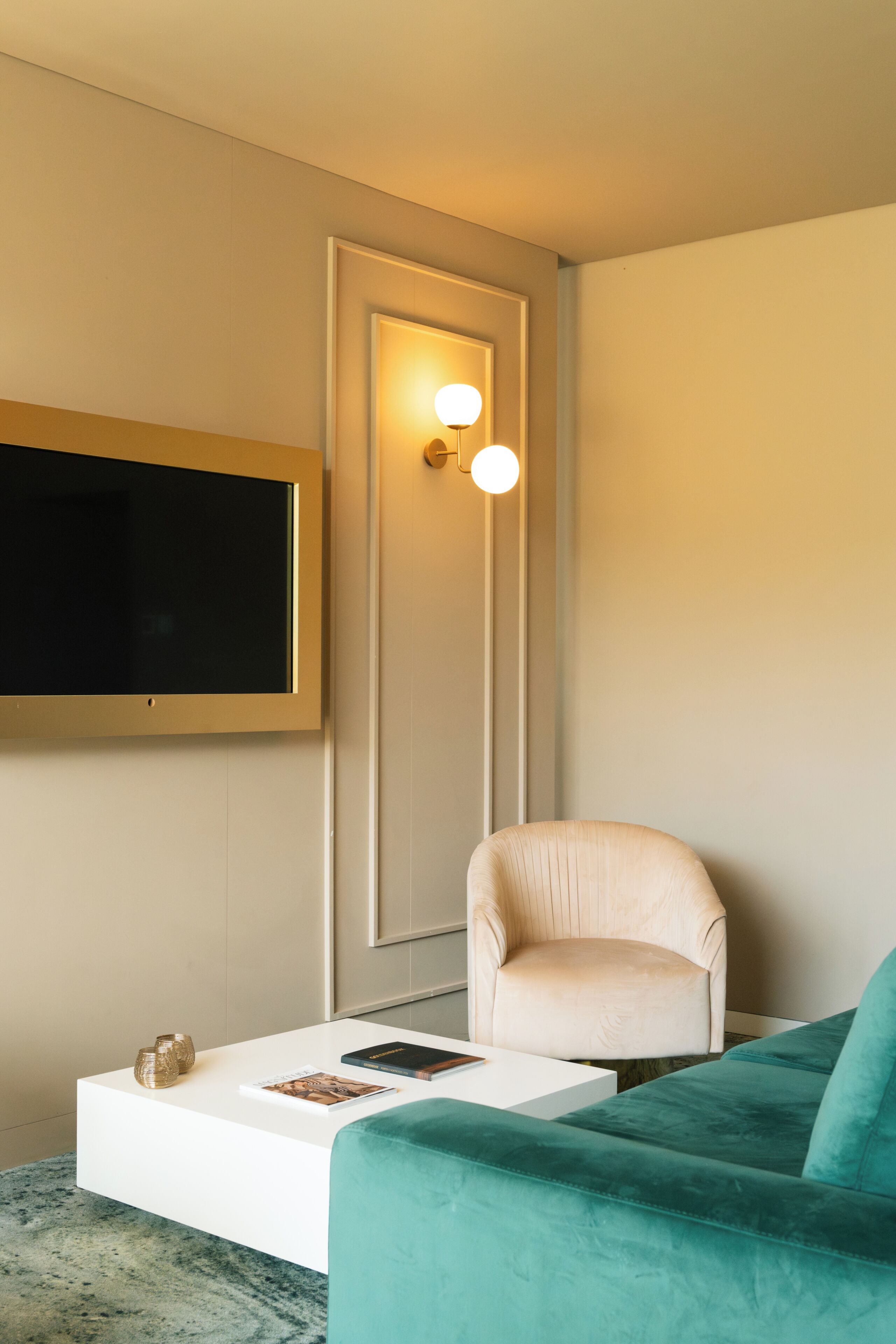 Quarto de hotel moderno com televisão de tela plana na parede, uma poltrona branca, um sofá verde-água e uma mesa de centro branca com uma revista.