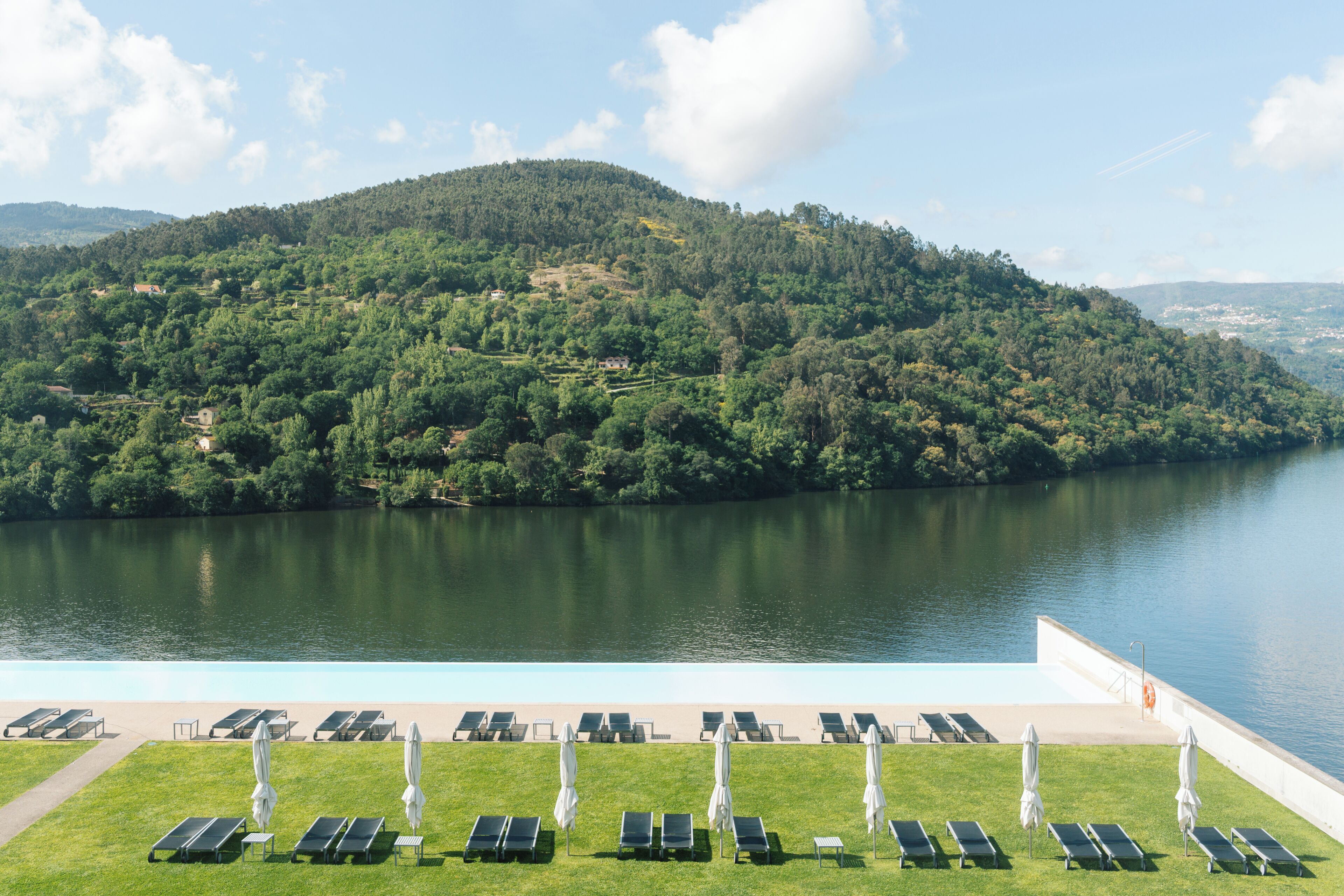 Vista para um lago com uma fileira de espreguiçadeiras e guarda-sóis, uma piscina e uma encosta ao fundo.