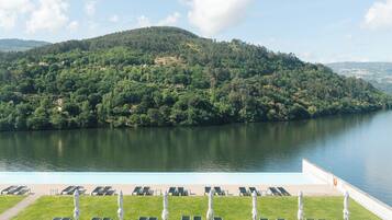 Vista para um lago com uma fileira de espreguiçadeiras e guarda-sóis, uma piscina e uma encosta ao fundo.