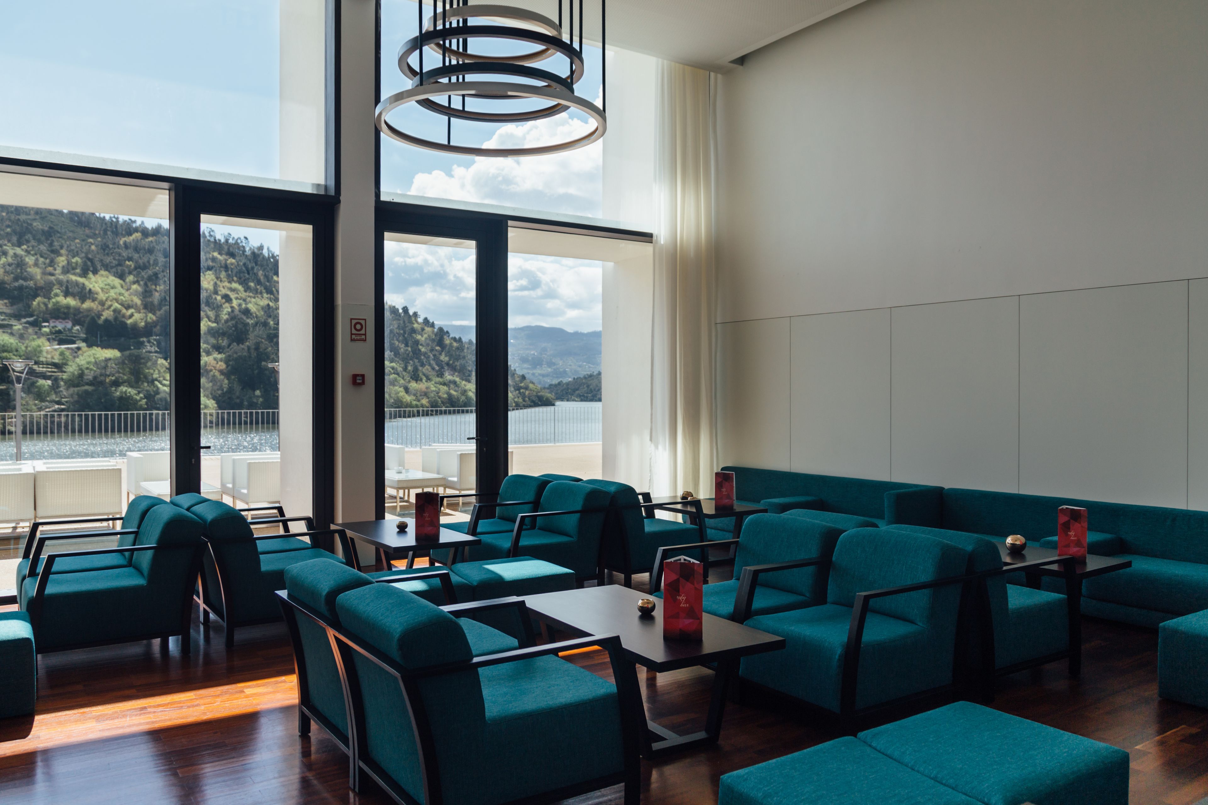 Uma área de estar moderna com assentos cor turquesa, grandes janelas com vista para uma paisagem montanhosa e uma luminária pendente contemporânea.