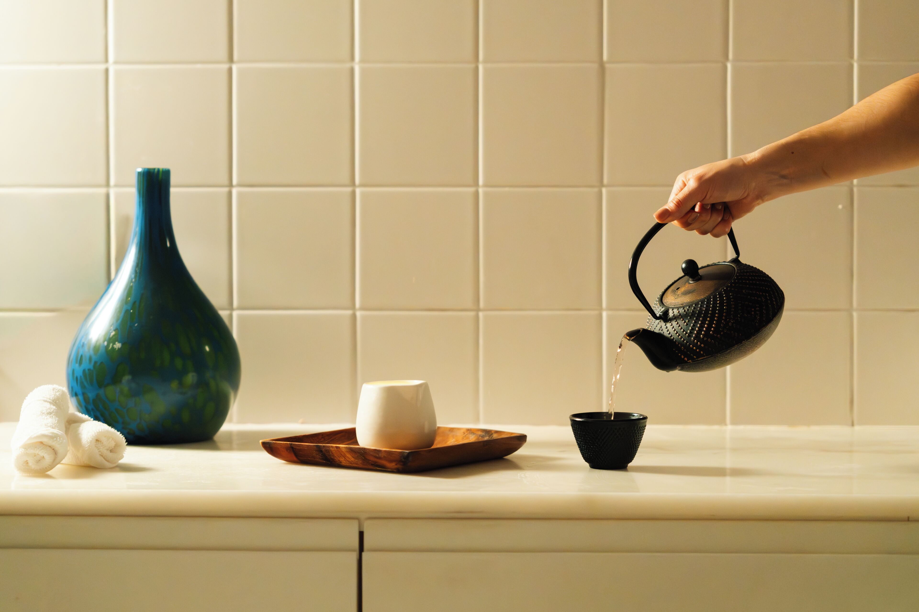 Uma mão despejando chá de um bule para uma xícara sobre uma bancada de cozinha, com um vaso e toalhas enroladas.