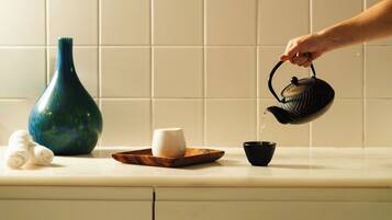 Uma mão despejando chá de um bule para uma xícara sobre uma bancada de cozinha, com um vaso e toalhas enroladas.