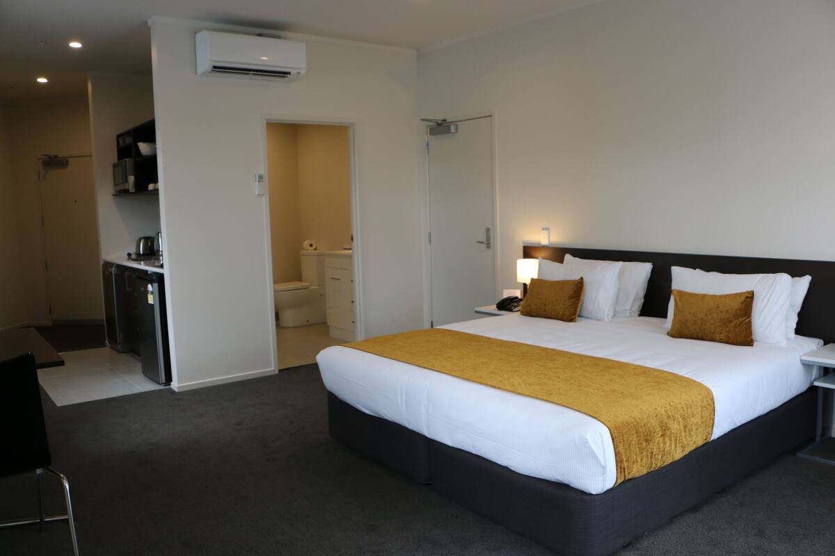 Quarto de hotel com uma cama grande, uma escrivaninha, uma cadeira, micro-ondas e uma pequena cozinha compacta.