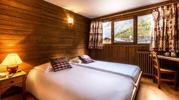 Quarto com paredes de madeira, cama com lençóis brancos, mesa de cabeceira com luminária, uma cadeira e uma janela com cortinas.