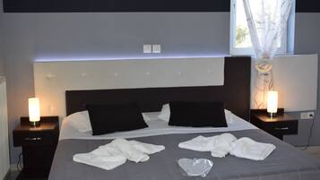Cama bem arrumada com roupa de cama branca, duas mesas de cabeceira com abajures e uma janela com cortinas finas.