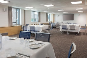 Sala de conferências com mesas preparadas para uma reunião, equipada com cadeiras, uma tela de projetor e uma grande janela que proporciona luz natural.