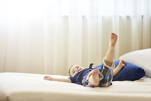 Uma criança deitada em uma cama com travesseiro azul, em um quarto com janela e cortinas.