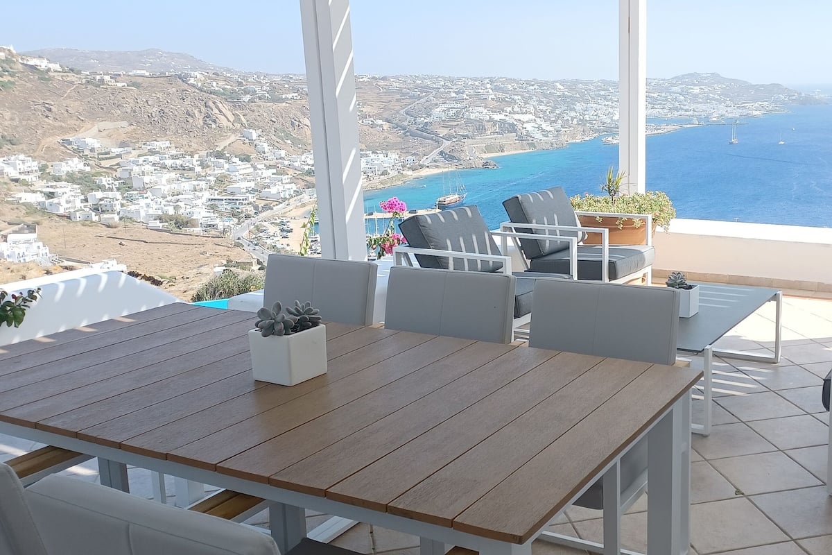 Uma varanda com mesa e cadeiras de madeira, com vista para uma cidade costeira e o mar.