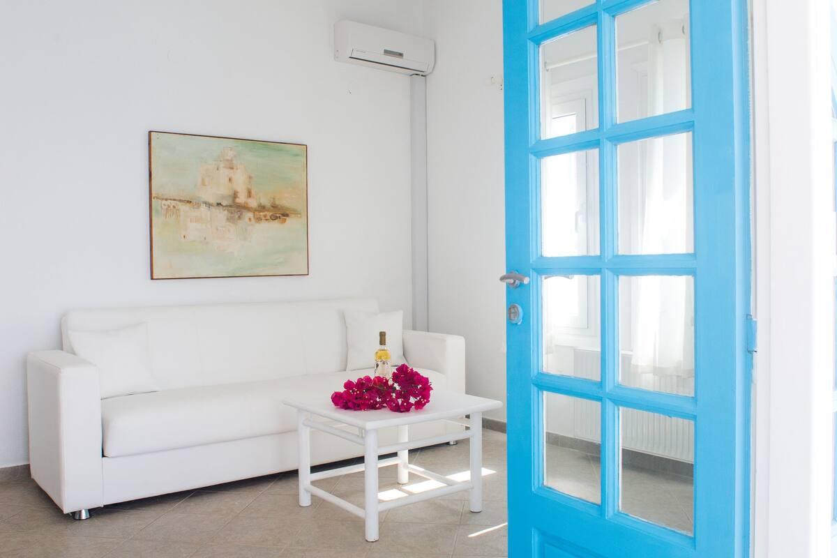 Quarto com sofá branco, uma mesinha pequena com um vaso de flores, porta azul e luminária pendente.
