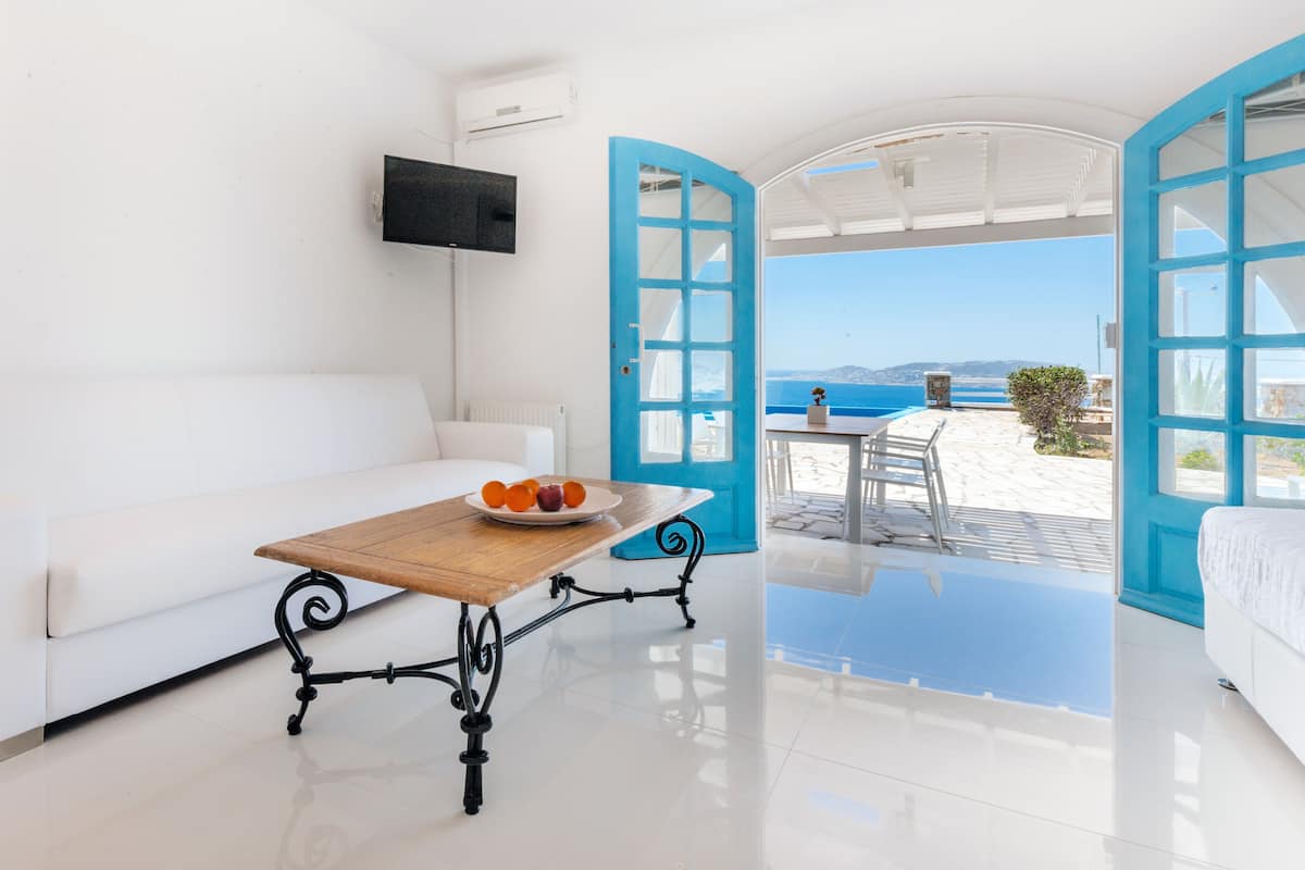Um quarto claro e arejado, com uma mesa de madeira, um sofá branco e uma TV fixada na parede. Há duas portas abertas que dão para uma varanda com vista para o mar.