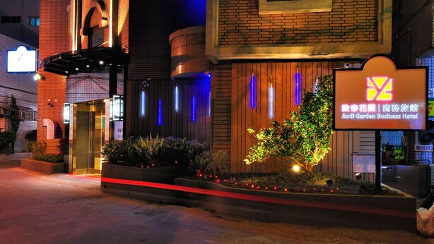 Entrada de hotel com placa, fachada de madeira e um pequeno jardim.