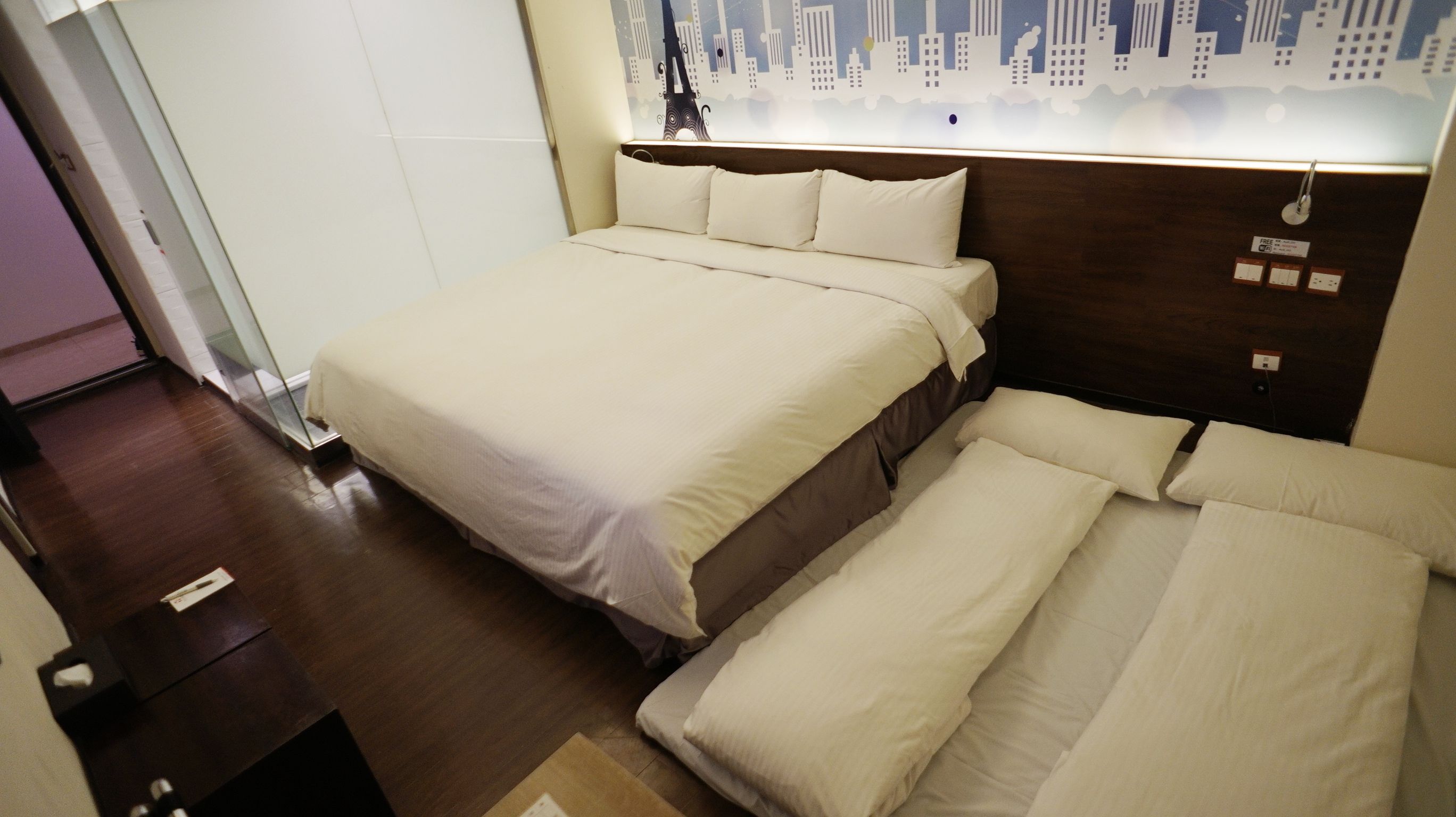 Quarto de hotel com uma cama grande, uma cama menor e uma mesinha de cabeceira.