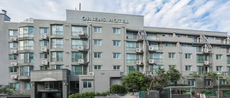 Um hotel de vários andares com um destaque para o letreiro "ORIENS HOTEL", várias varandas e uma entrada com portão.