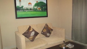 Sala de estar com um sofá bege, duas almofadas estampadas, um quadro de um edifício e uma mesa de centro de madeira escura com um copo e um objeto de metal.