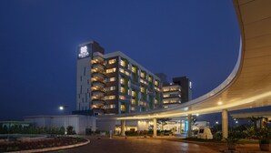 Um hotel com vários andares e design moderno, com uma entrada destacada, uma cobertura e uma área ajardinada na frente.
