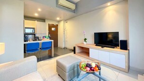 Uma sala de estar moderna com uma televisão de tela plana, uma mesa de centro de vidro com frutas, um sofá e uma área de cozinha com cadeiras azuis.