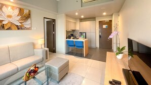 Uma sala de estar moderna com um sofá, um banquinho e uma mesa de centro de vidro com frutas. Uma televisão e um controle remoto estão sobre uma mesa de madeira. Uma cozinha com cadeiras azuis é visível ao fundo.