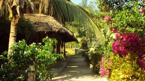 Um caminho leva a uma construção com telhado de palha, cercada por vegetação exuberante e plantas floridas.