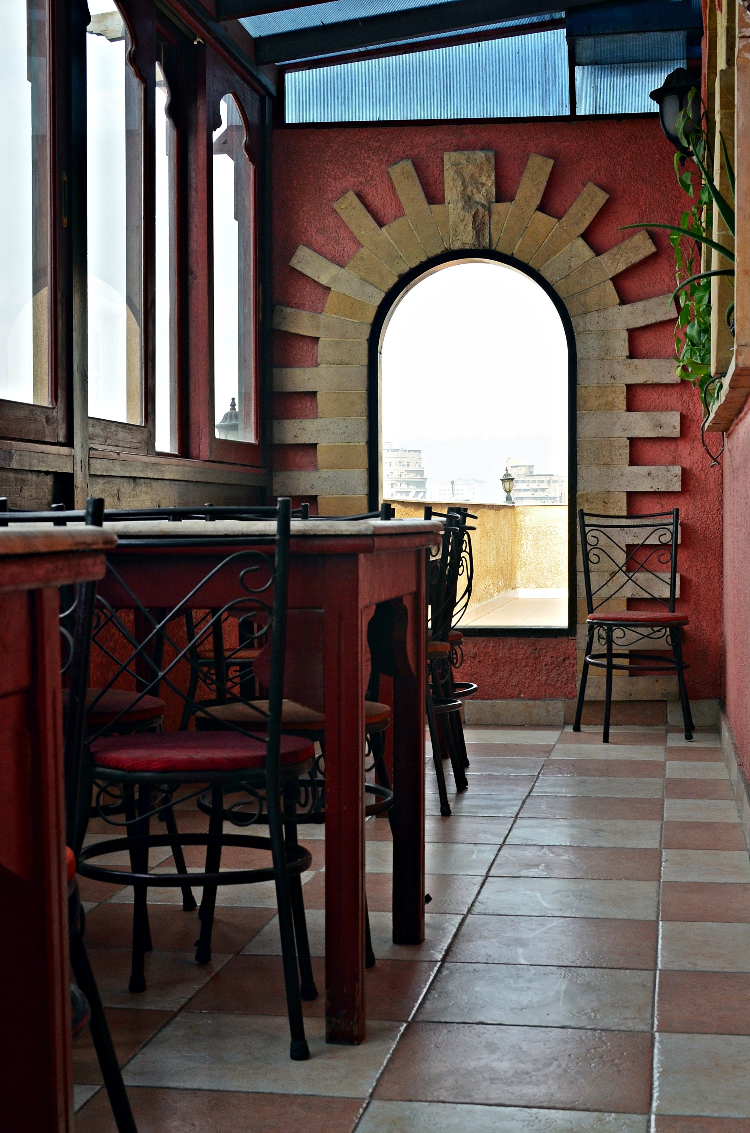 Um restaurante com vista através de uma janela em arco, com chão em azulejos e mobiliário de madeira.