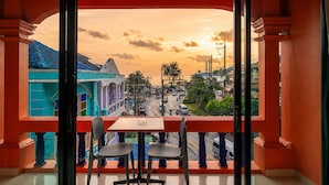 Varanda com mesa e duas cadeiras, vista para uma rua com prédios e um pôr do sol.
