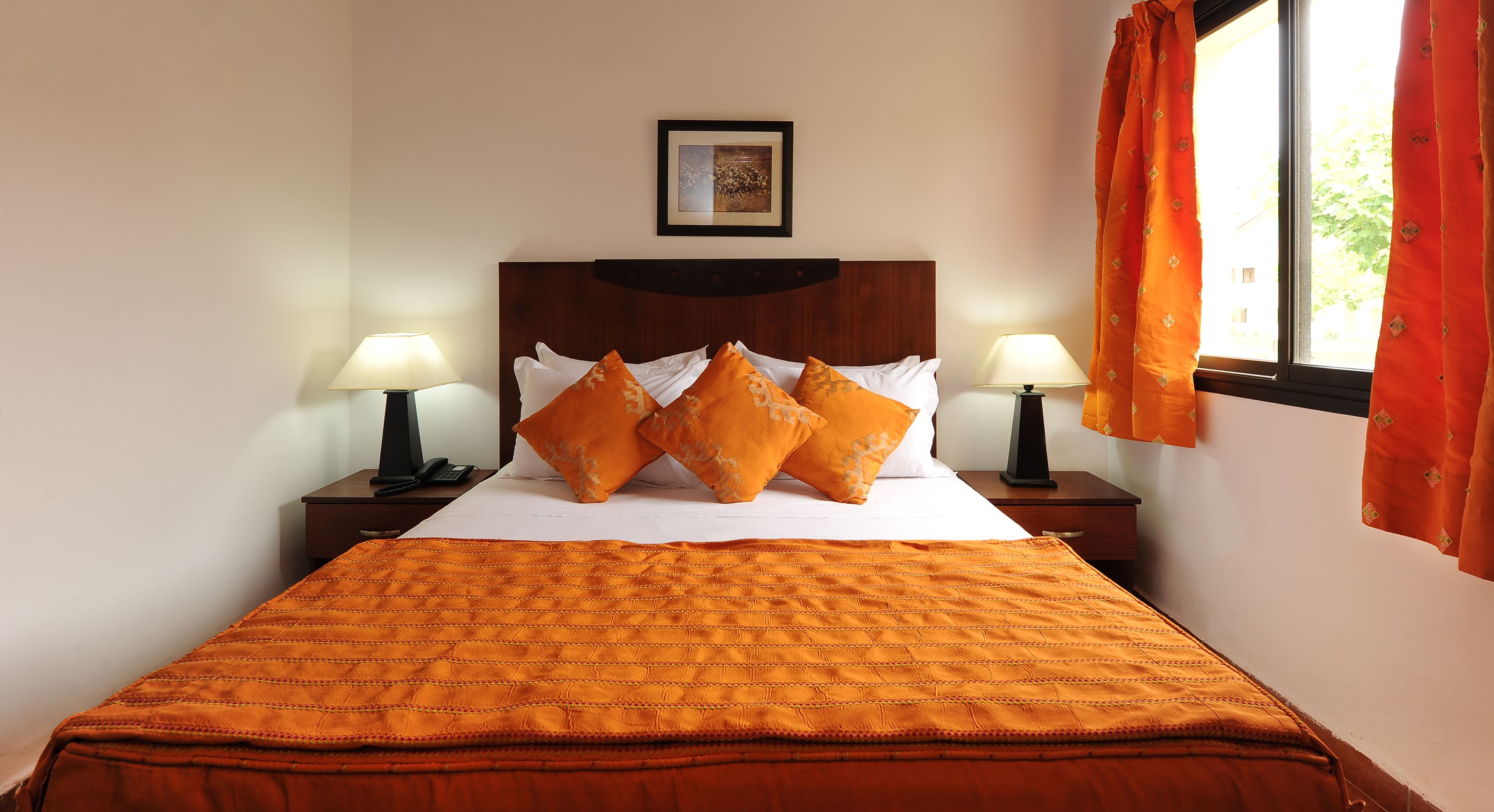 Cama bem arrumada com colcha e almofadas laranja, duas luminárias de cabeceira, um quadro na parede e uma janela com cortinas laranja.
