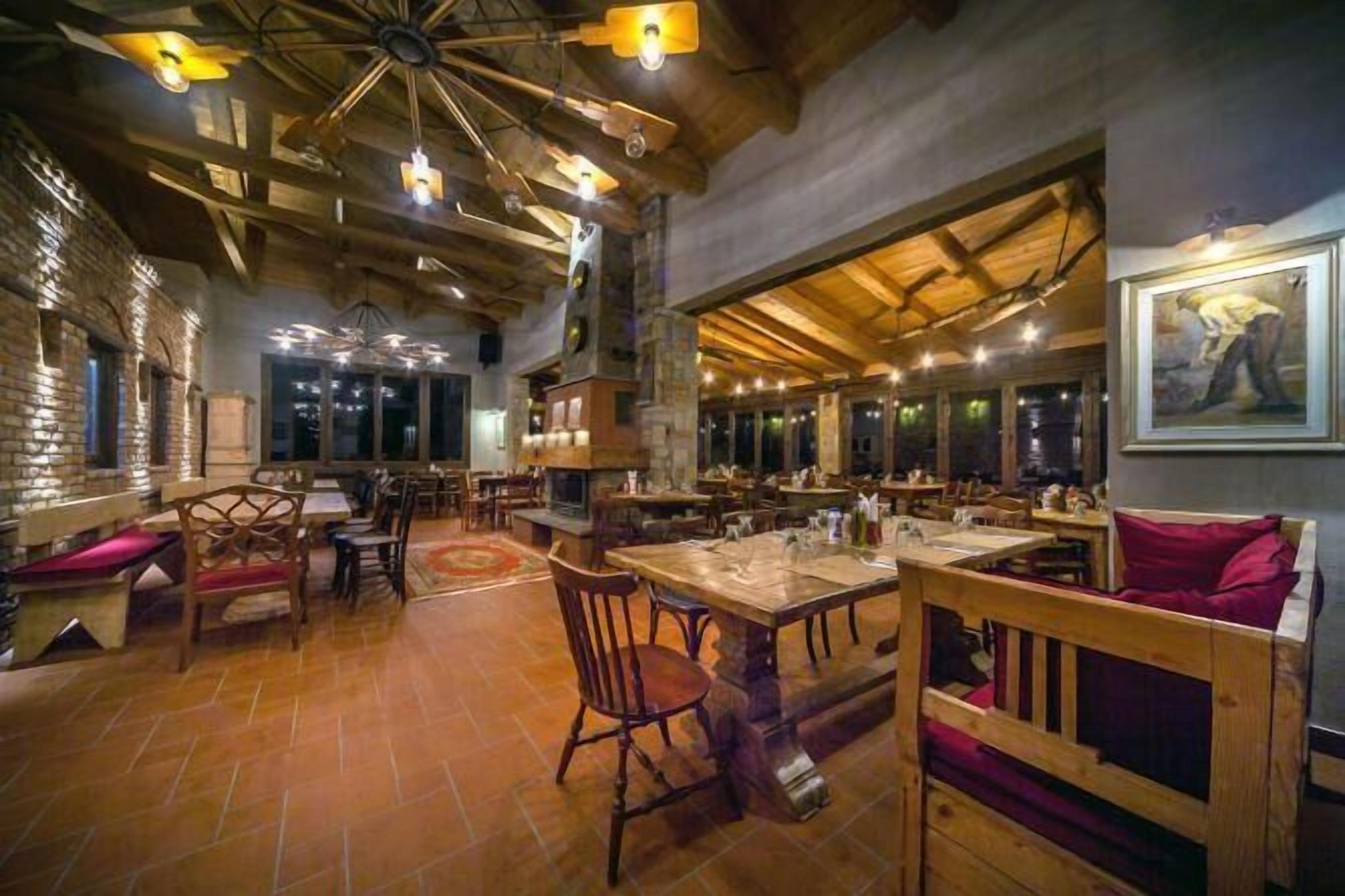 Um restaurante espaçoso com pé direito alto, vigas aparentes e ambiente rústico. Mesas e cadeiras dispostas para refeições, e uma lareira visível ao fundo.
