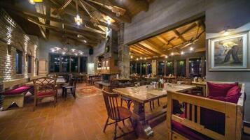Um restaurante espaçoso com pé direito alto, vigas aparentes e ambiente rústico. Mesas e cadeiras dispostas para refeições, e uma lareira visível ao fundo.