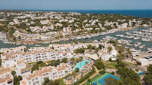 Vista aérea de uma cidade costeira com edifícios brancos, um porto com vários barcos e uma grande piscina rodeada por espreguiçadeiras e palmeiras.