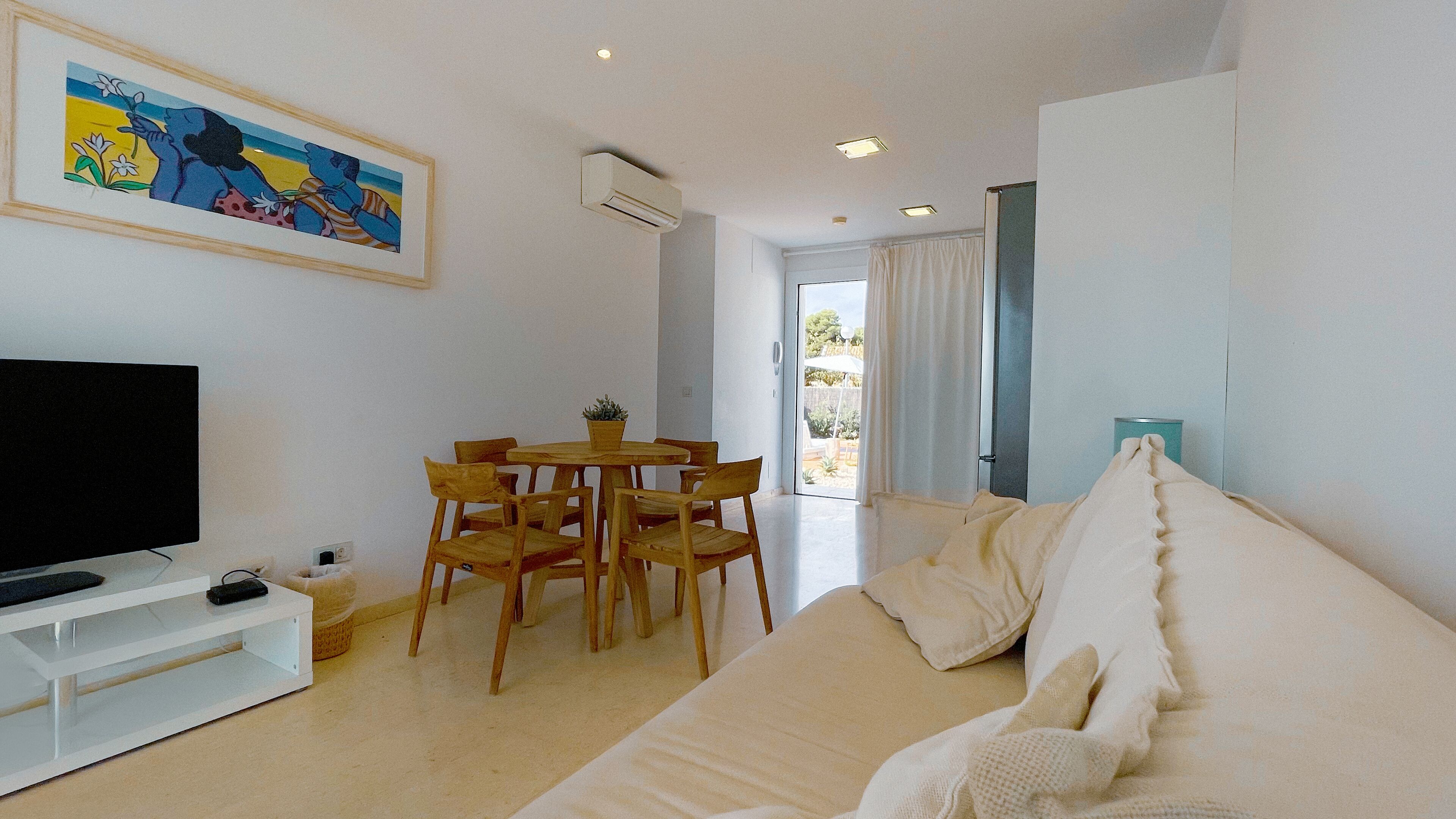 Uma sala de estar com televisão, mesa de jantar de madeira, cadeiras, sofá e um quadro na parede.