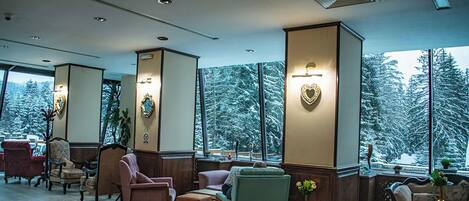 Um hall amplo com várias áreas de estar, grandes janelas com vista para árvores cobertas de neve e uma lareira com um espelho decorativo.
