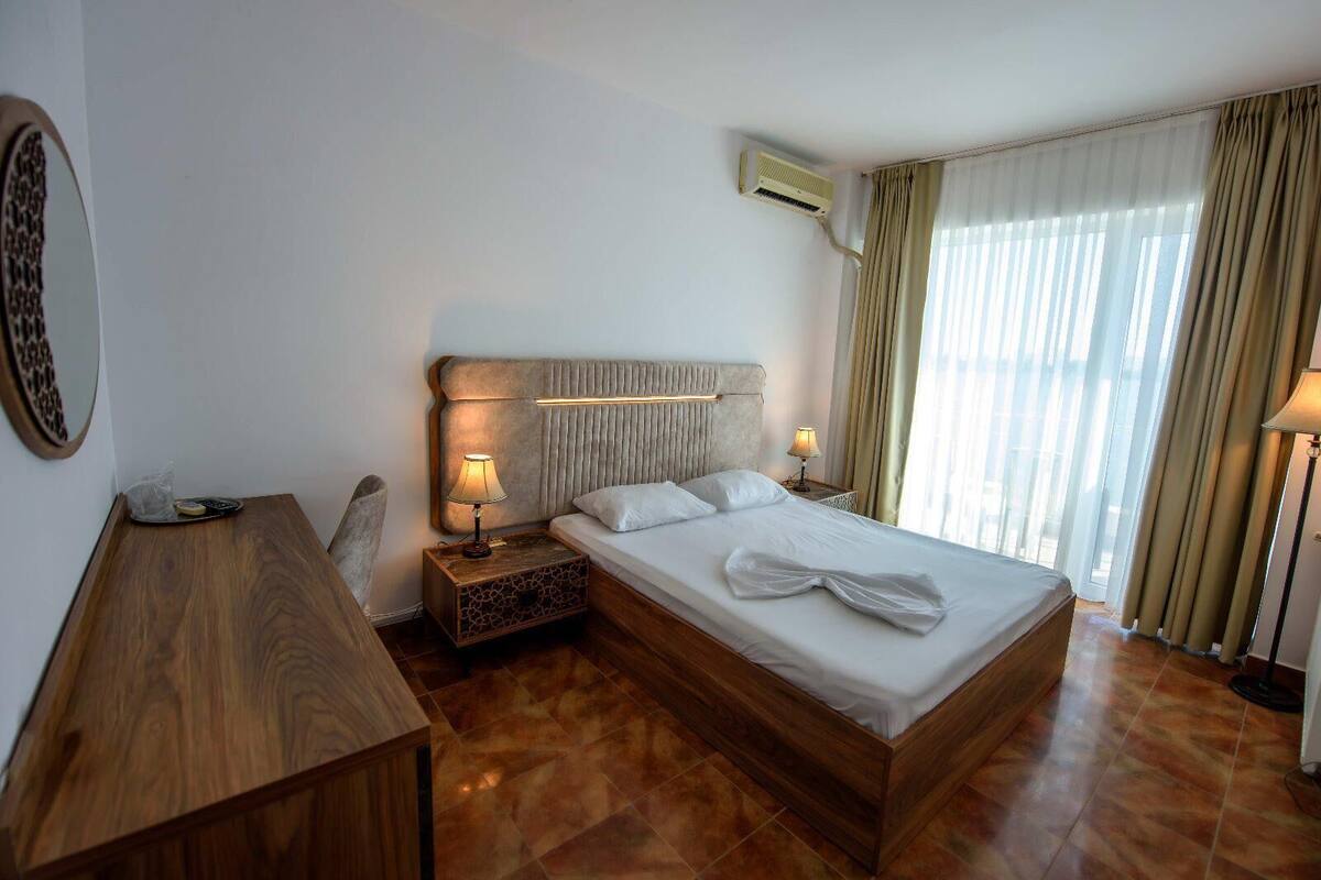 Quarto de hotel com uma cama grande, duas mesas de cabeceira com luminárias, uma escrivaninha de madeira e um ar condicionado fixado na parede.
