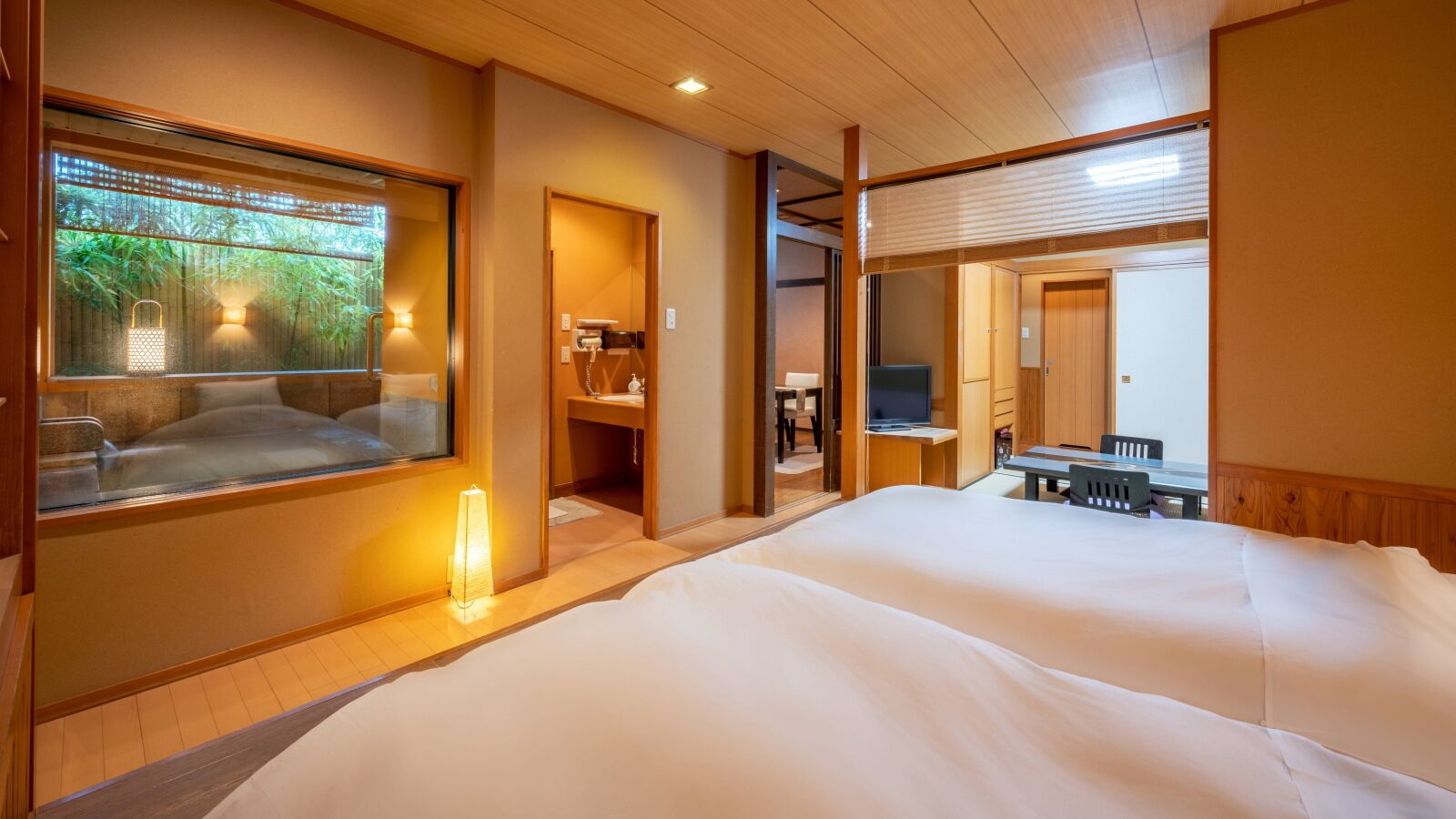 Quarto de hotel com uma cama grande, uma mesa com cadeira, televisão e banheiro com pia e espelho.