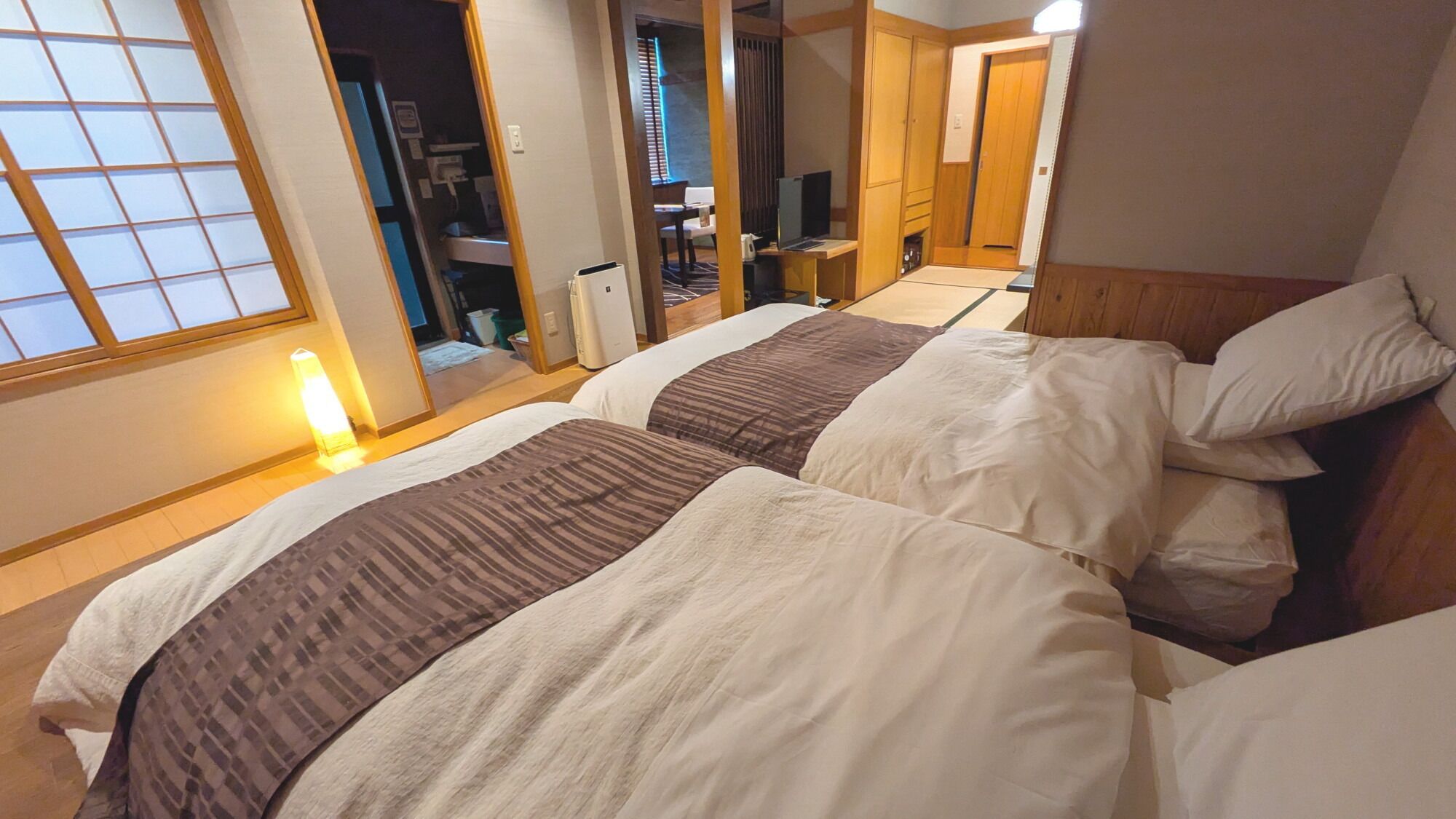 Quarto de hotel com duas camas, cabeceira de madeira, uma pequena mesa de trabalho com cadeira e vista através de uma porta de correr.