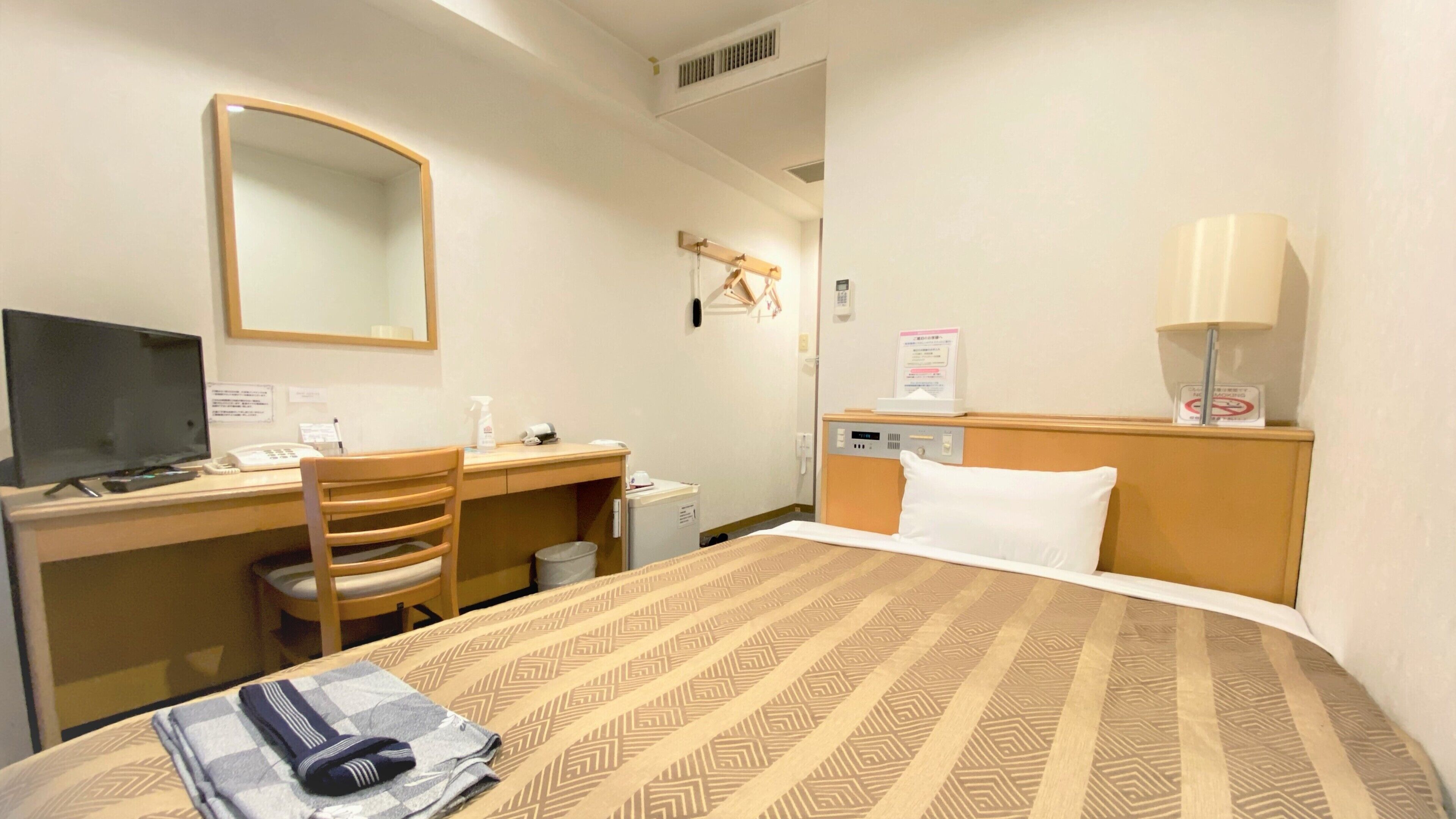 Quarto de hotel com cama, escrivaninha, cadeira, televisão e espelho.