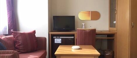 Quarto de hotel pequeno com uma escrivaninha de madeira, televisão, armário e sofá.