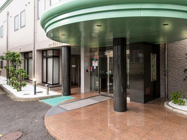 Entrada de hotel com toldo de vidro curvo, porta de metal e pequena área externa com um banco.