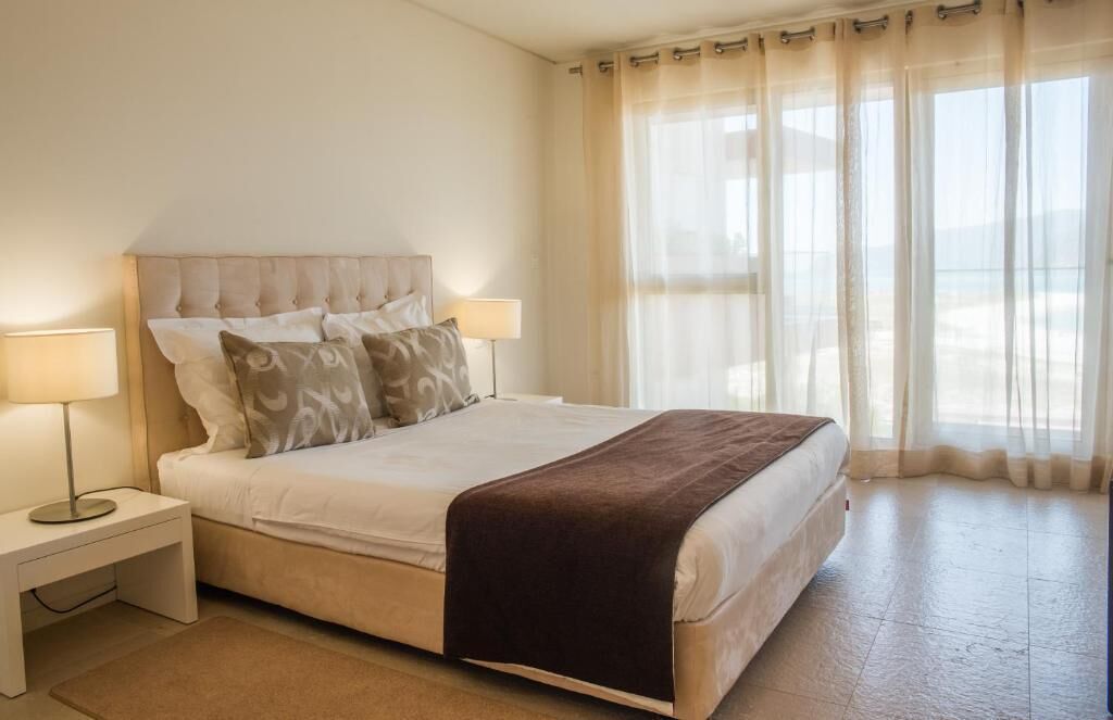 Cama bem arrumada com manta marrom, duas luminárias de cabeceira e uma janela com cortinas leves.
