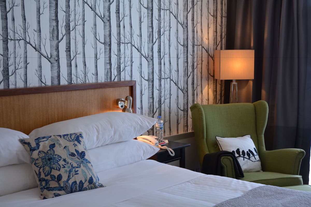 Quarto de hotel com cama, mesa de cabeceira com abajur, poltrona verde e parede com padrão de árvore.