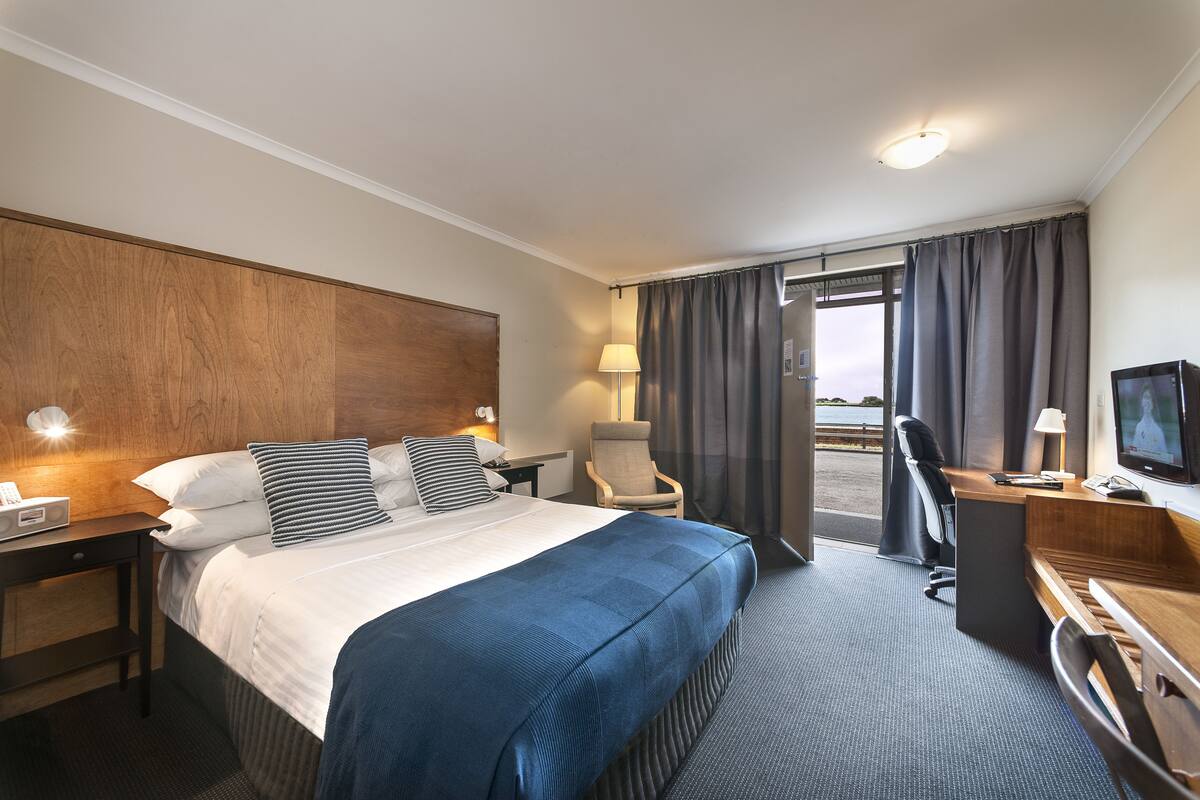 Quarto de hotel com uma cama grande, escrivaninha, cadeira, televisão e janela com cortinas.