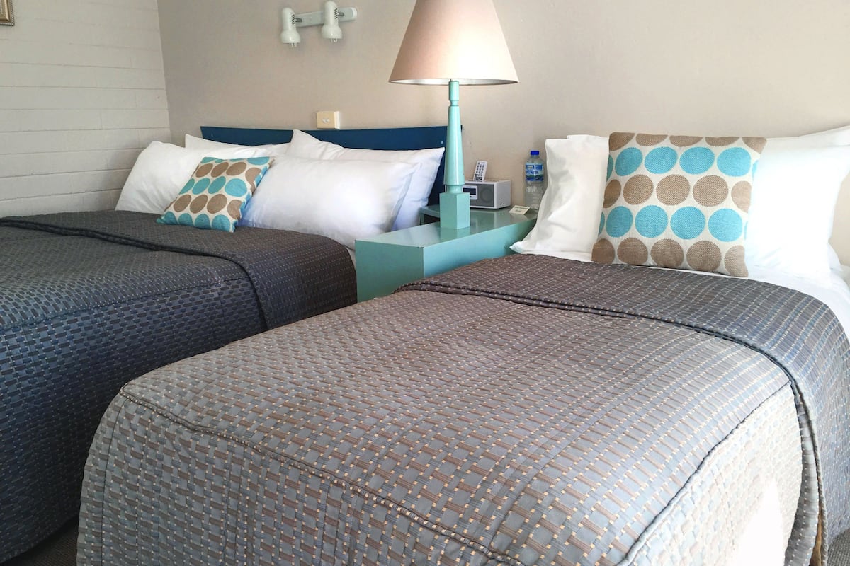 Quarto de hotel com duas camas, uma mesinha de cabeceira, um abajur e uma tomada na parede.