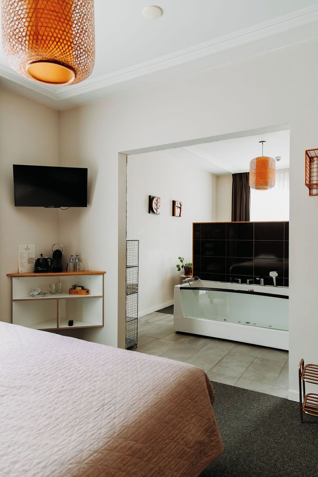 Quarto de hotel moderno com cama, TV, banheiro com box de vidro e área de refeições com prateleira.